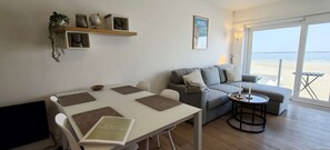Uma sala de estar moderna com um sofá, uma mesa de jantar com cadeiras, uma pequena mesa redonda com uma vela e vista para a praia através de grandes janelas.