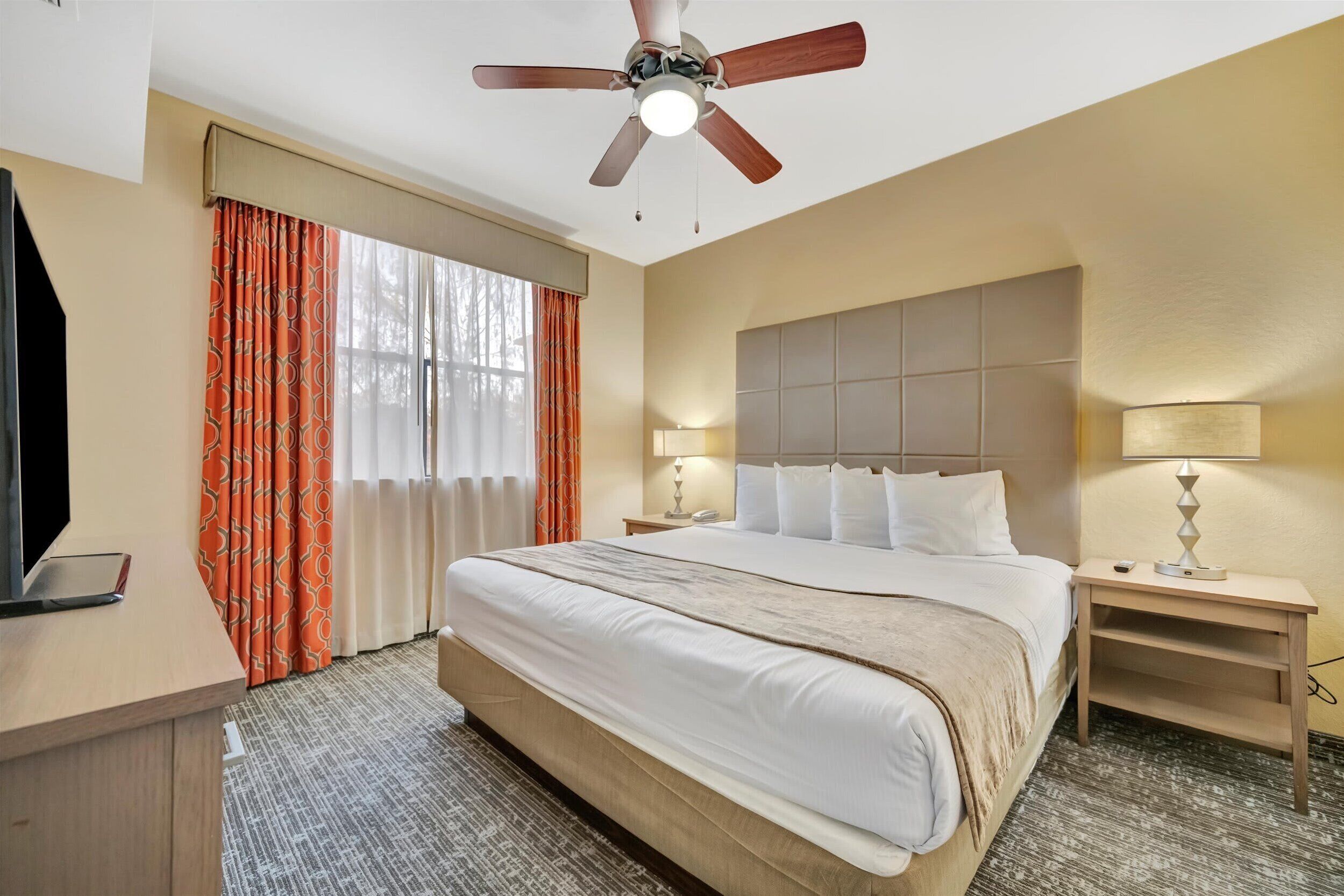 Quarto de hotel com uma cama grande, duas mesas de cabeceira com luminárias, um ventilador de teto, uma televisão e uma janela com cortinas.