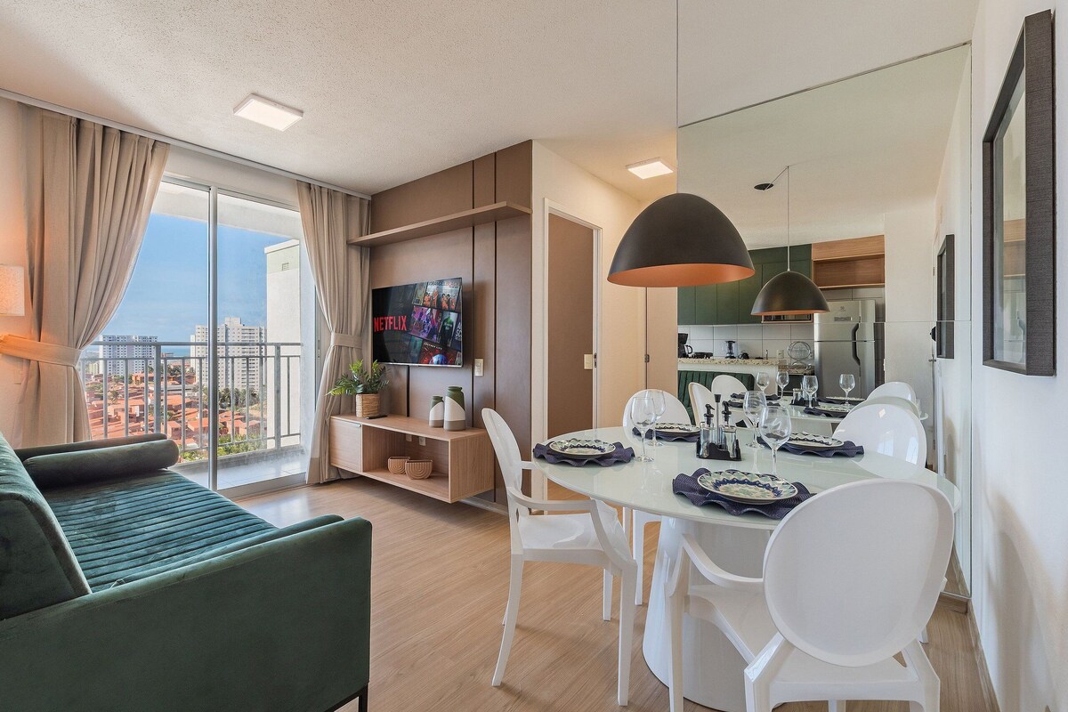 Uma sala de estar moderna com um sofá verde, uma mesa de jantar posta para quatro pessoas e uma cozinha ao fundo.