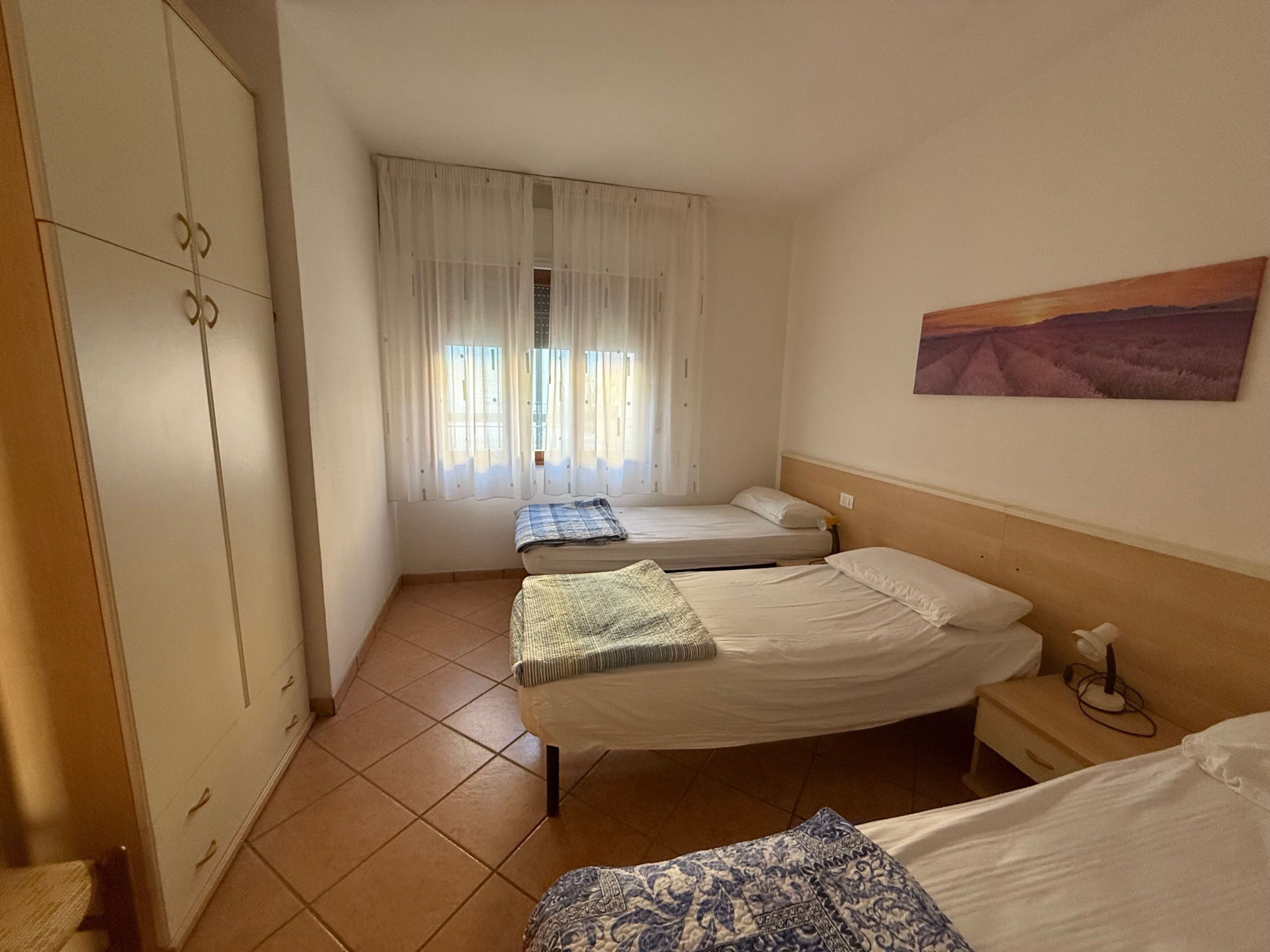 Quarto de hotel com duas camas de solteiro, um guarda-roupa, uma janela com cortinas e um quadro na parede.