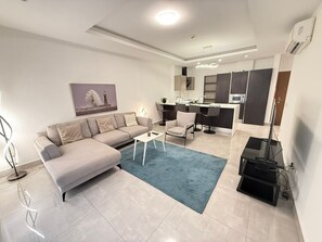 Uma sala de estar moderna com sofá, mesa de centro e tapete azul. Há uma área de cozinha com armários e micro-ondas.