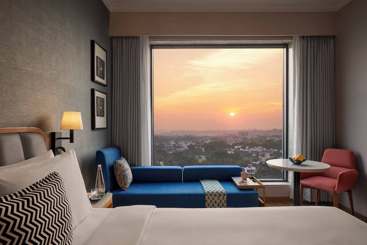 Quarto de hotel com uma janela grande, uma cama, um sofá azul, uma cadeira vermelha e uma mesinha com uma tigela de frutas.