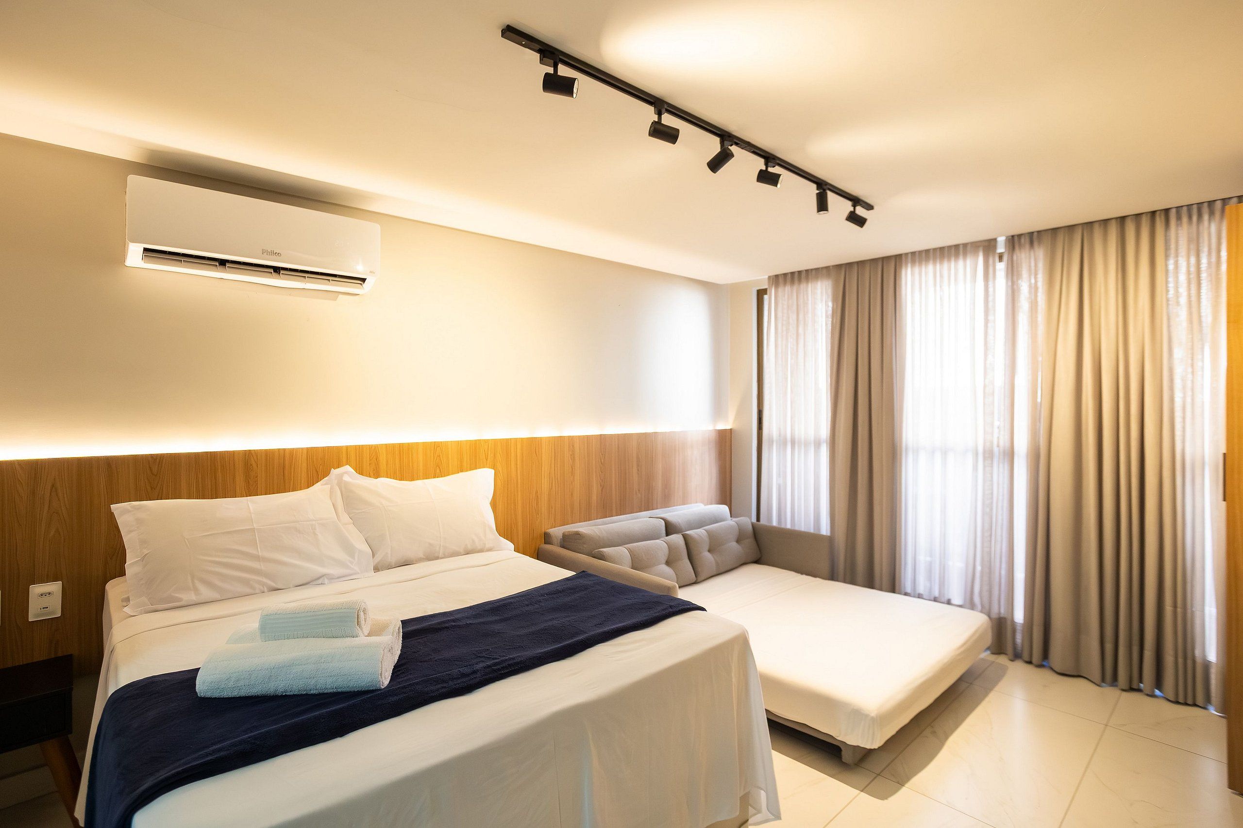 Quarto de hotel com cama, sofá, ar-condicionado e janela com cortinas.