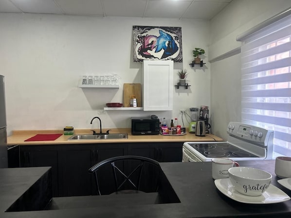 Cozinha compacta com micro-ondas, fogão e pia. Há uma mesa de jantar com cadeiras e uma mesa de centro. Um quadro está pendurado na parede acima da pia.