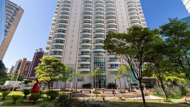 Um hotel moderno e alto, com entrada central, rodeado por jardins bem cuidados e palmeiras.