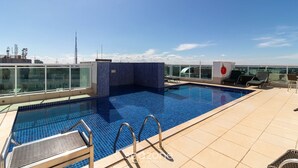 Uma piscina no terraço com azulejos azuis vibrantes, guarda-corpo de vidro e vista para o horizonte da cidade.