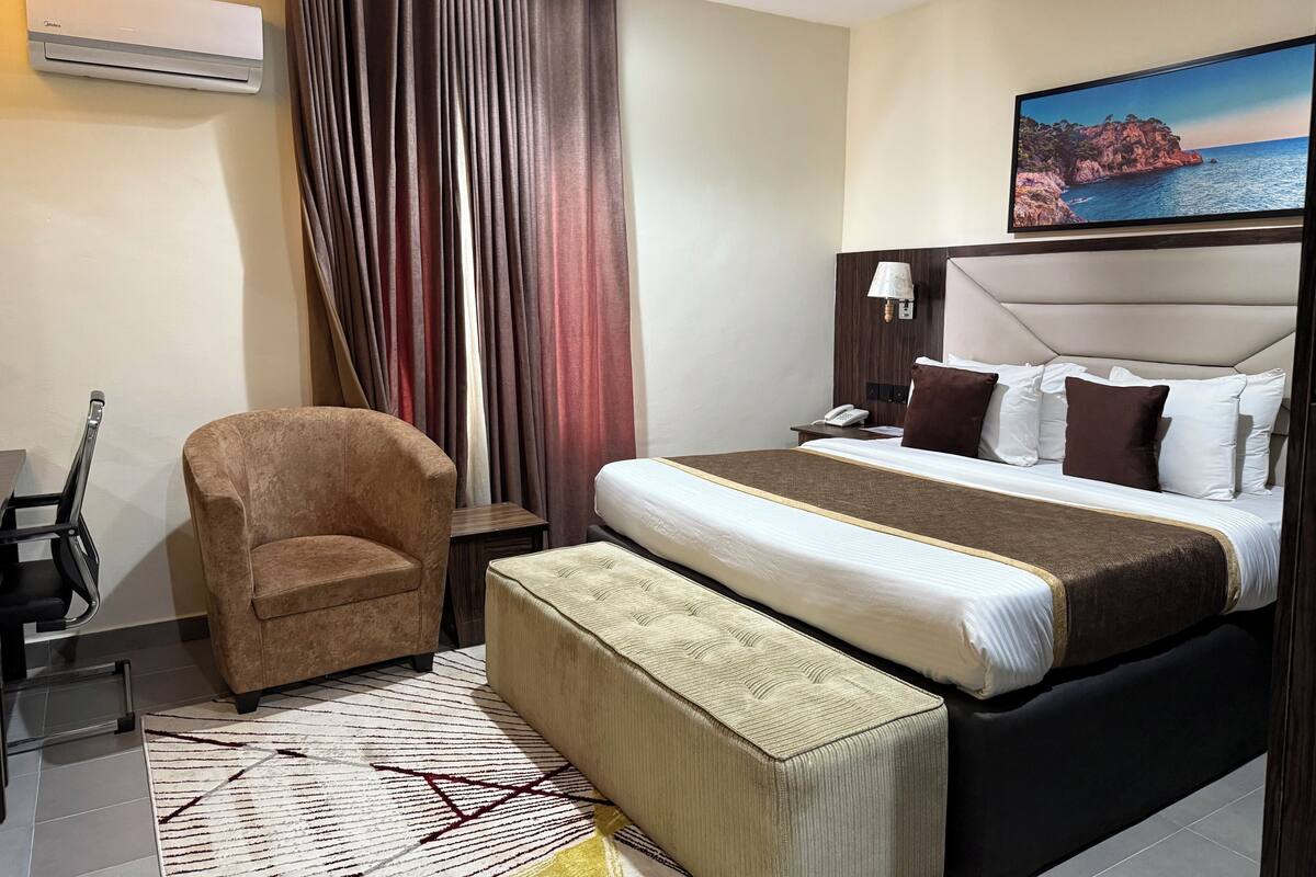 Quarto de hotel com cama, poltrona, escrivaninha, ar condicionado e pintura de uma paisagem costeira.