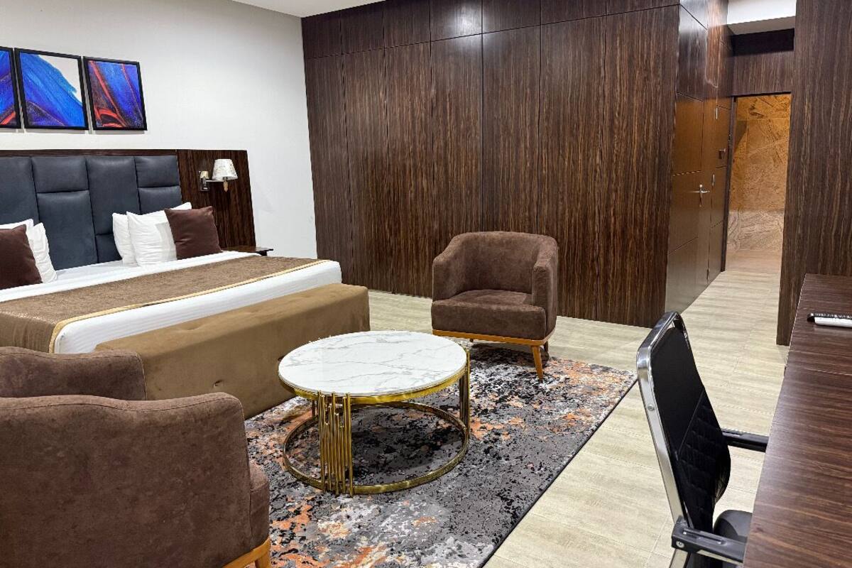 Quarto de hotel moderno com uma cama grande, duas poltronas, uma mesa de centro redonda e uma parede de madeira escura em destaque.