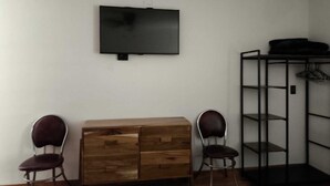 Quarto com cômoda de madeira, duas cadeiras, uma televisão e estante de metal.