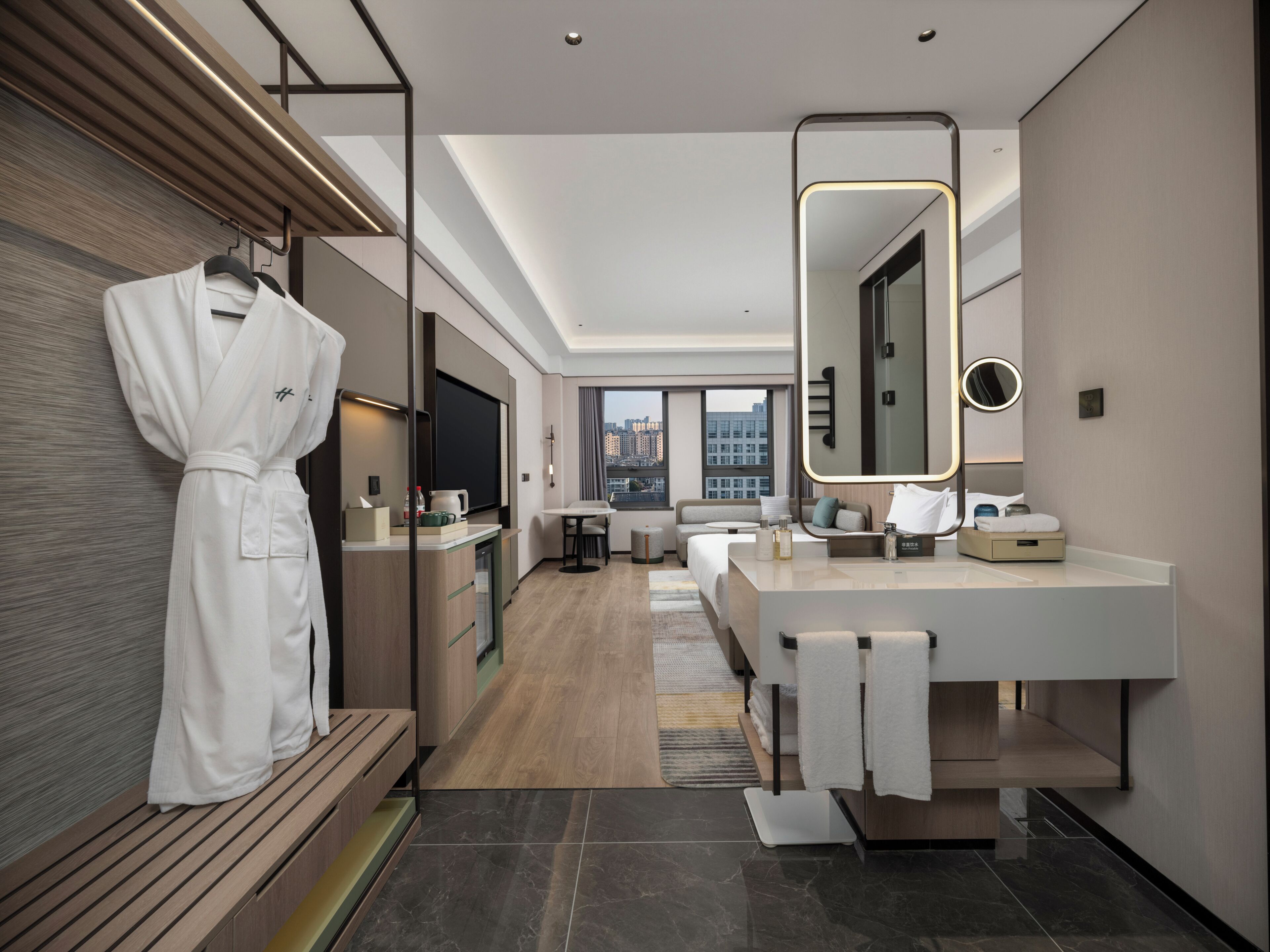Quarto de hotel moderno com um espelho grande, um roupão pendurado em um porta-roupas, uma secretária com cadeira, um sofá e vista da cidade através de janelas amplas.
