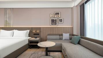 Quarto de hotel moderno com cama grande, sofá, mesa pequena e mesa de cabeceira. Há quadros nas paredes e uma janela com cortinas.