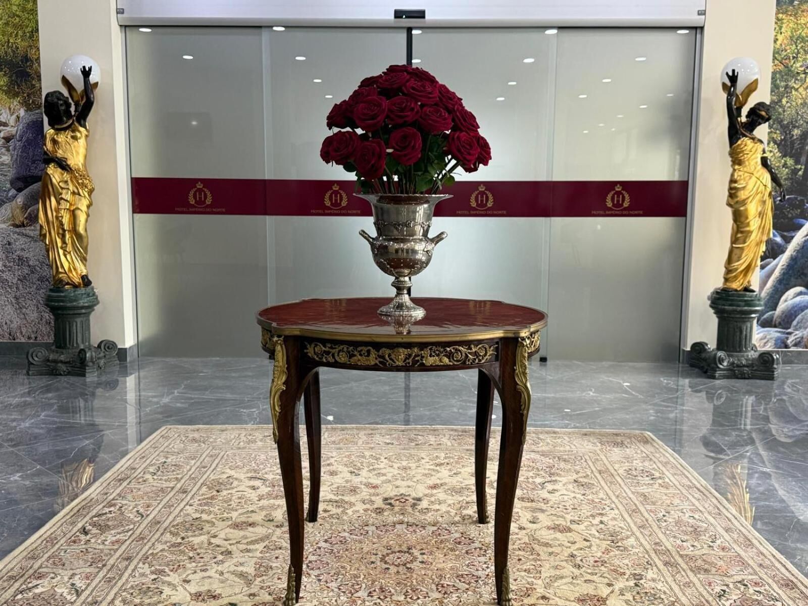 Um hall com uma mesa redonda, um vaso de rosas vermelhas e duas estátuas douradas.