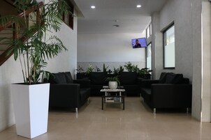 Um hall moderno com sofás pretos, uma mesa de centro, plantas em vasos e uma tela de televisão.