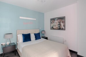 Quarto com cama, duas mesas de cabeceira, um criado-mudo, prateleira na parede e uma moldura com imagem.