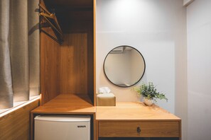 Quarto de hotel com guarda-roupa de madeira, um espelho redondo, uma pequena mesa de cabeceira com um porta-lenços e uma planta em vaso, e um mini-frigorífico embutido com a etiqueta 'WINIA'.