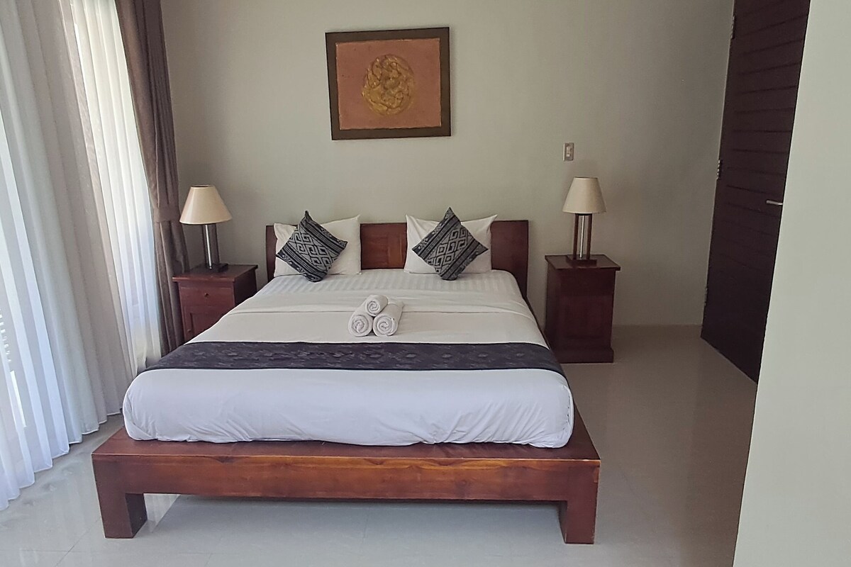 Quarto com cama, duas mesas de cabeceira, janela com cortinas e um quadro na parede.