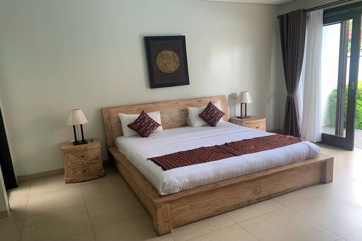 Quarto com cama de madeira, duas mesas de cabeceira, uma obra de arte na parede e vista através de uma porta de vidro.