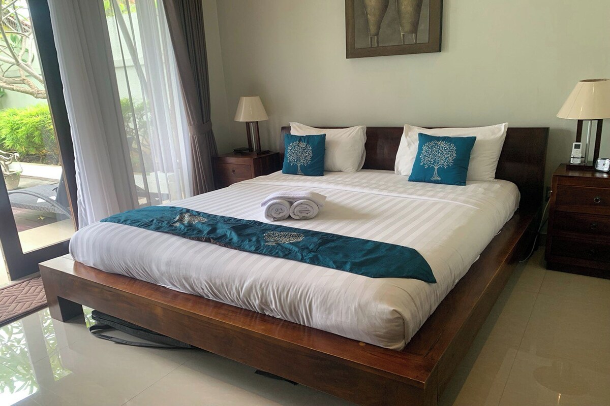Cama bem arrumada com colcha azul e branca, duas almofadas com desenhos de árvores e um par de toalhas enroladas.