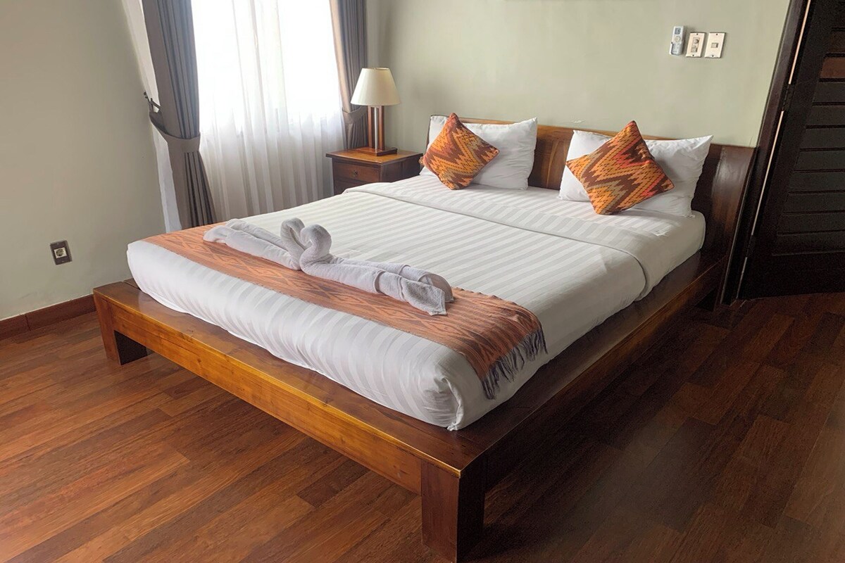 Uma cama bem arrumada com estrutura de madeira, uma mesa de cabeceira com abajur e uma janela com cortinas.