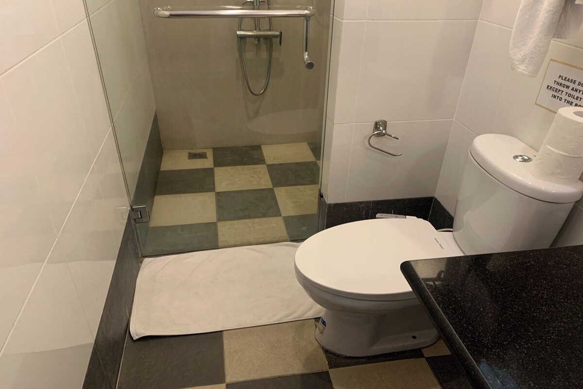 Um banheiro com vaso sanitário, chuveiro com barra de apoio e chão quadriculado.