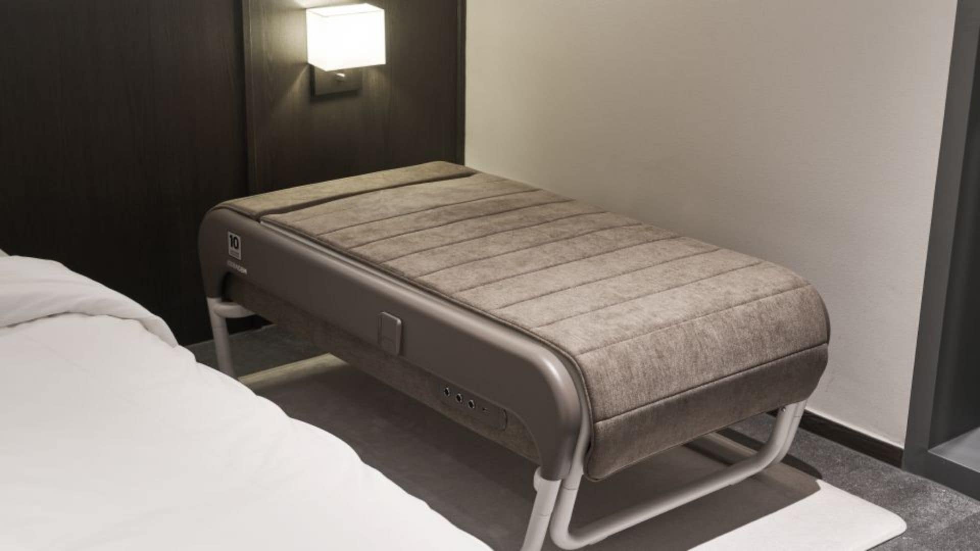 Uma cama compacta com cabeceira, mesa de cabeceira e luminária fixada na parede.