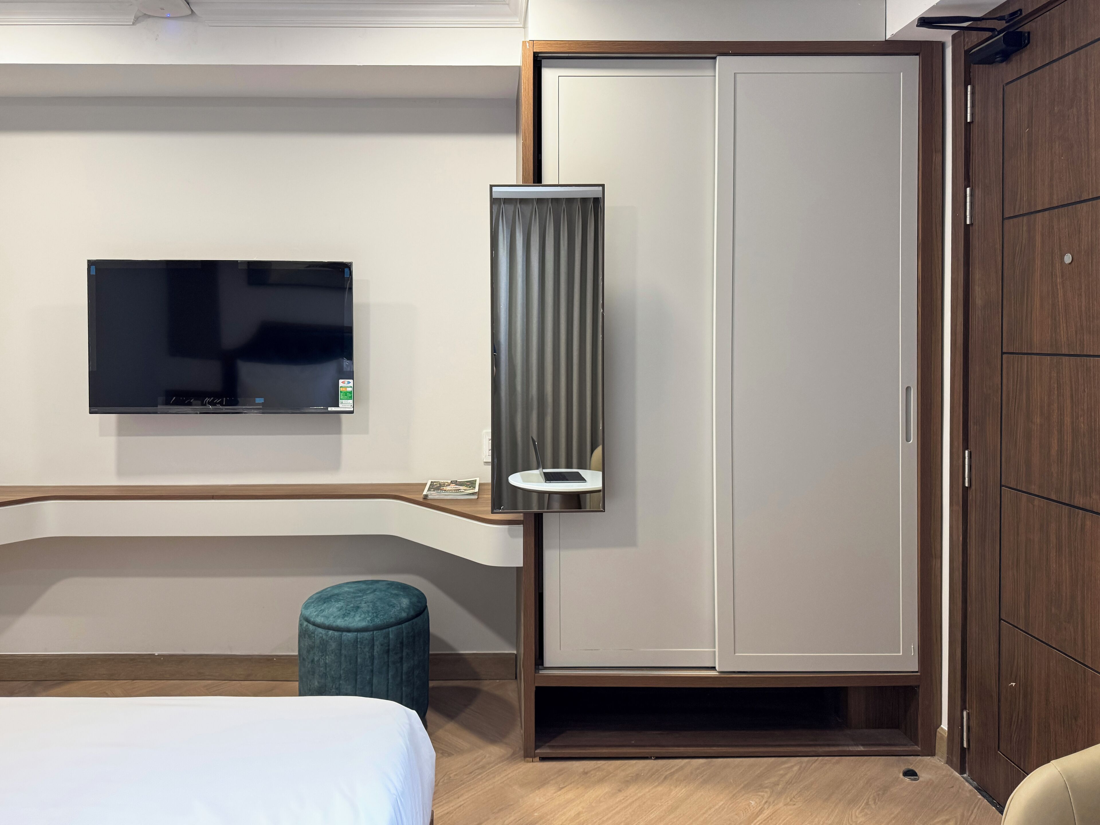 Quarto de hotel com cama, televisão na parede, guarda-roupa e uma pequena mesa com abajur.