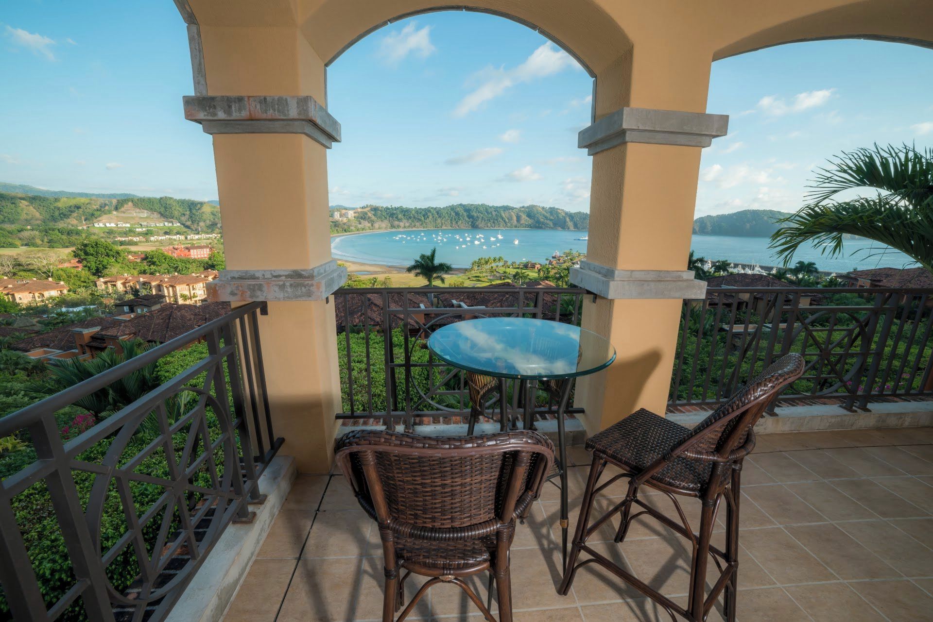 Varanda com mesa de vidro e cadeiras de vime, com vista para a paisagem costeira com edifícios e barcos.