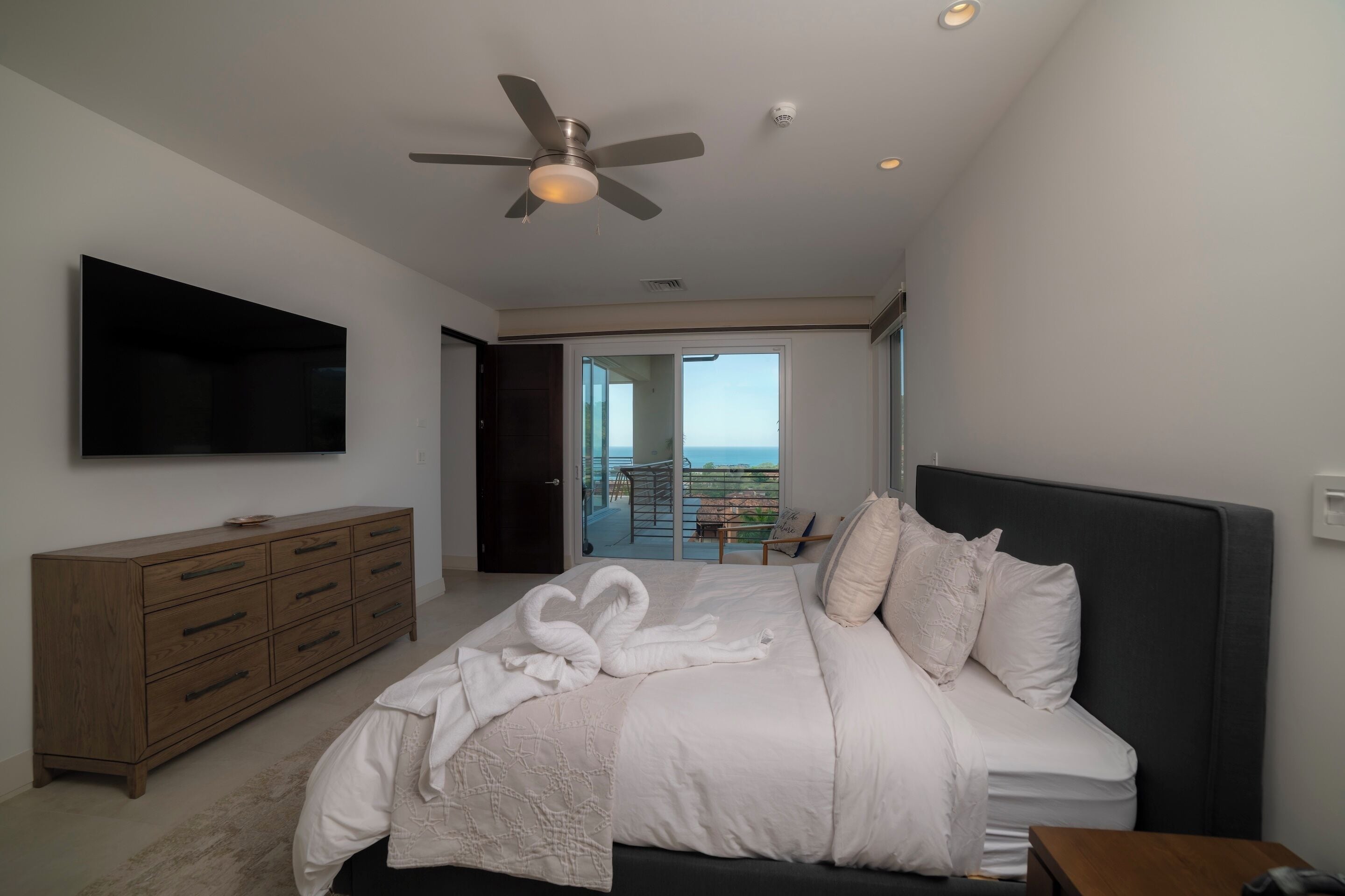 Quarto moderno com cama grande, ventilador de teto, TV de tela plana e vista para o exterior.