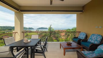 Uma varanda com mesa, cadeiras e um sofá, com vista para uma paisagem deslumbrante com colinas e um corpo de água.