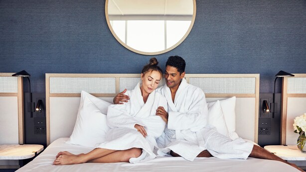 Um casal em roupões brancos sentado numa cama com cabeceira de madeira, um espelho redondo acima e mesas de cabeceira com abajures e um vaso de flores.