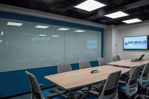 Sala de conferências com paredes de vidro, uma tela grande exibindo 'NEURALIS' e várias mesas com cadeiras.