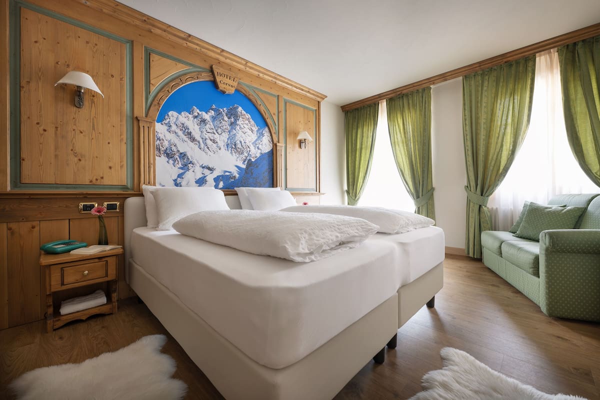 Quarto de hotel com uma cama grande, painéis de madeira, uma pintura de paisagem montanhosa, um sofá verde e uma mesa de cabeceira de madeira com um vaso e um livro.