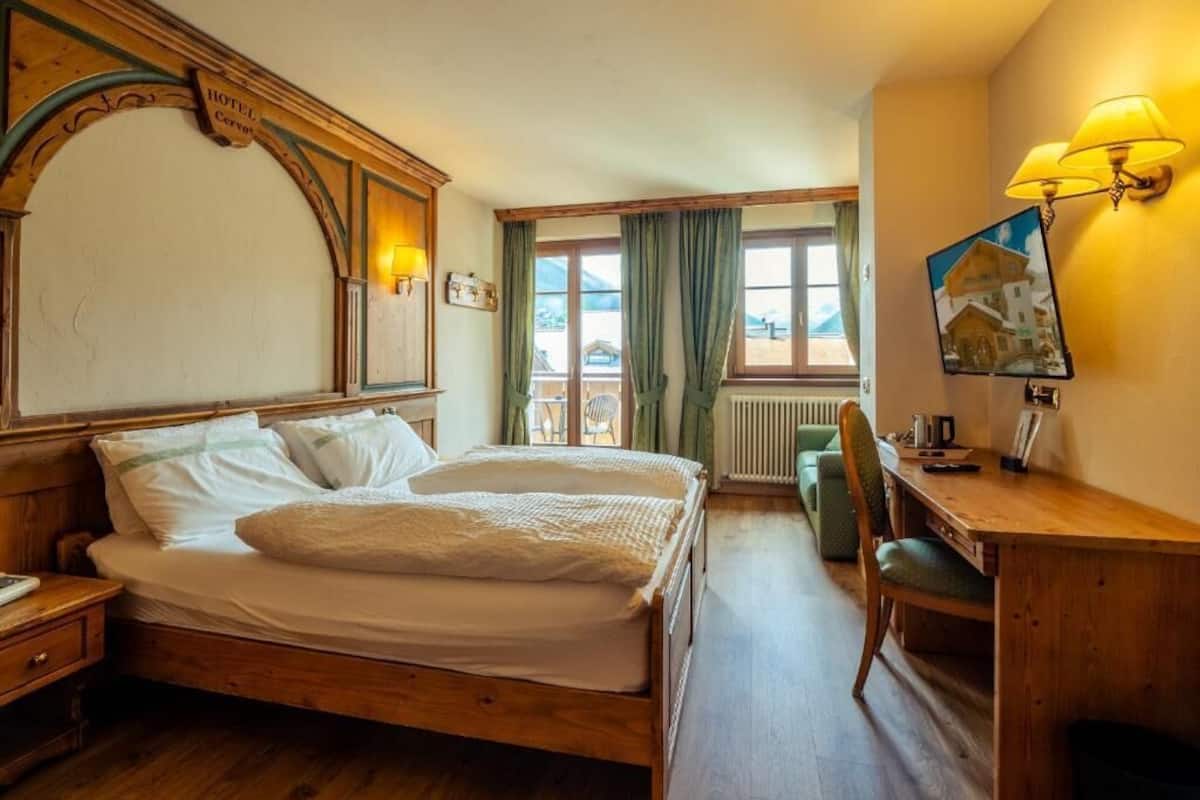 Quarto de hotel com cama de madeira, uma escrivaninha com luminária, uma cadeira e vista para as montanhas pela janela.
