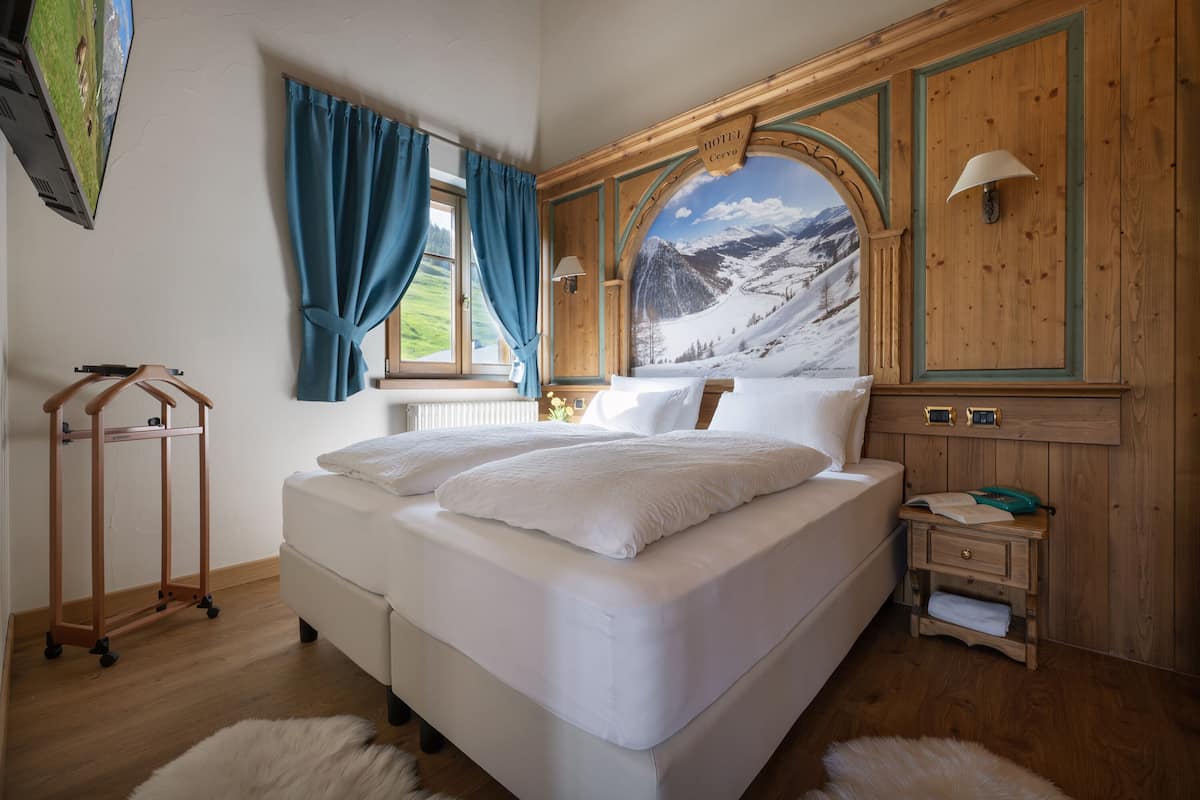 Quarto com cama grande, painéis de madeira, janela com cortinas, luminária na parede e mesa de cabeceira pequena.
