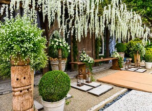 Um jardim com flores brancas pendentes, vasos de plantas, um banco de madeira e um caminho de pedra.