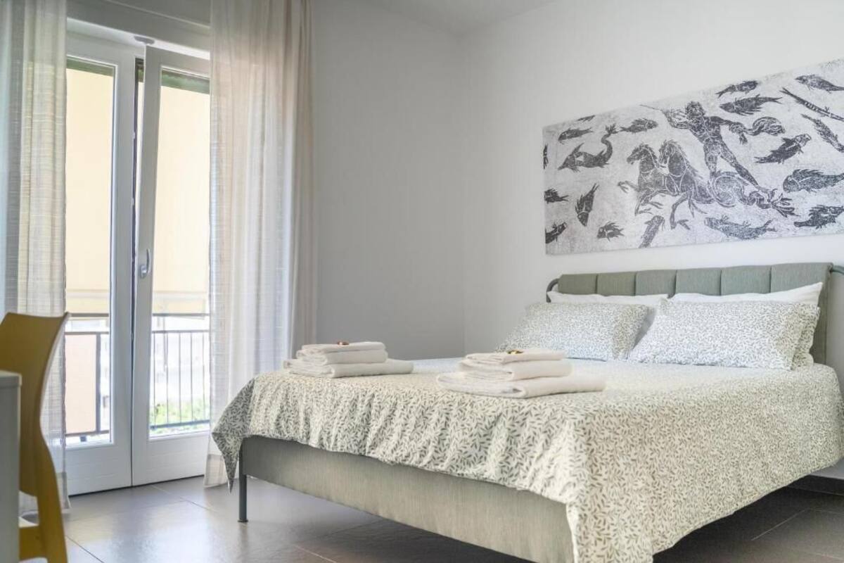 Quarto com cama, cadeira, varanda com cortinas e uma obra de arte na parede.