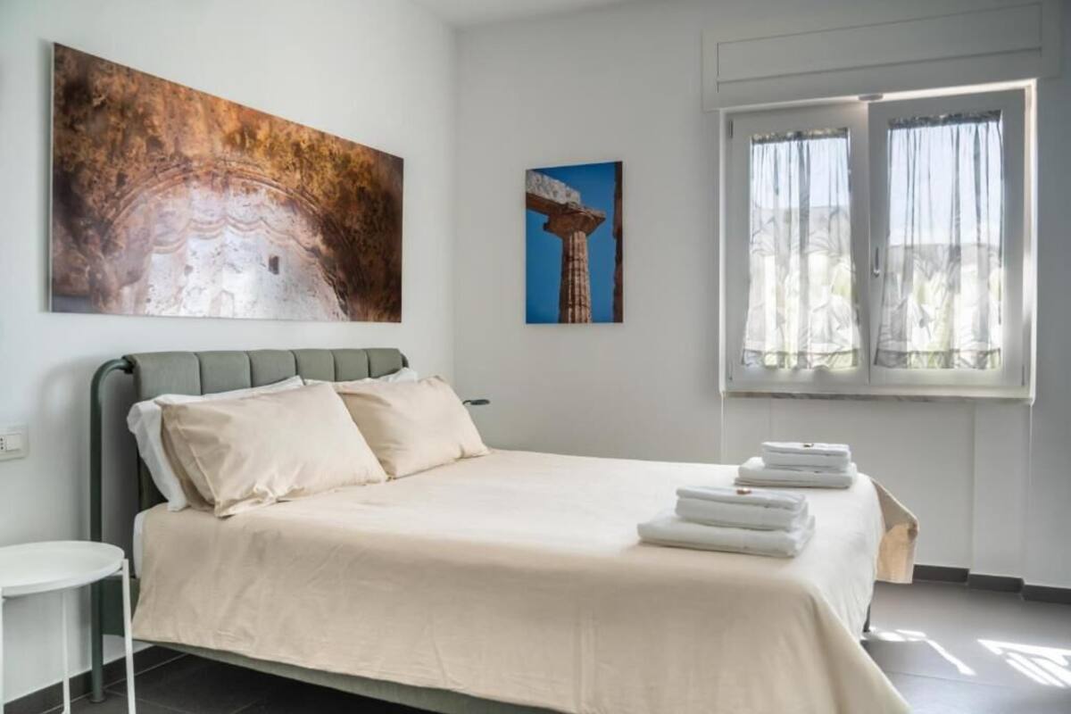 Um quarto com uma cama, uma mesinha de cabeceira, uma janela com cortinas e duas obras de arte penduradas na parede.