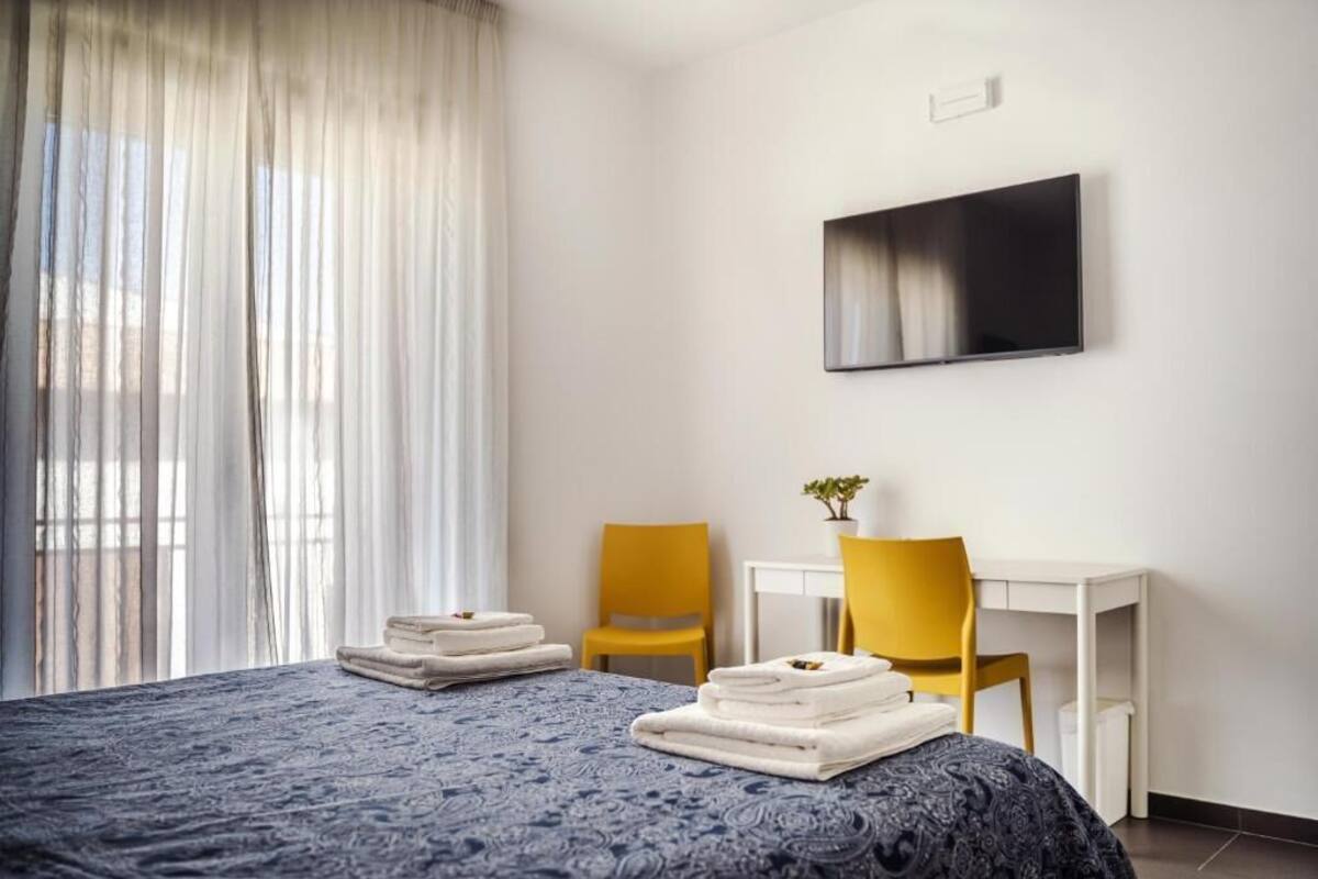 Quarto de hotel com cama, escrivaninha, cadeira, televisão e janela com cortinas.