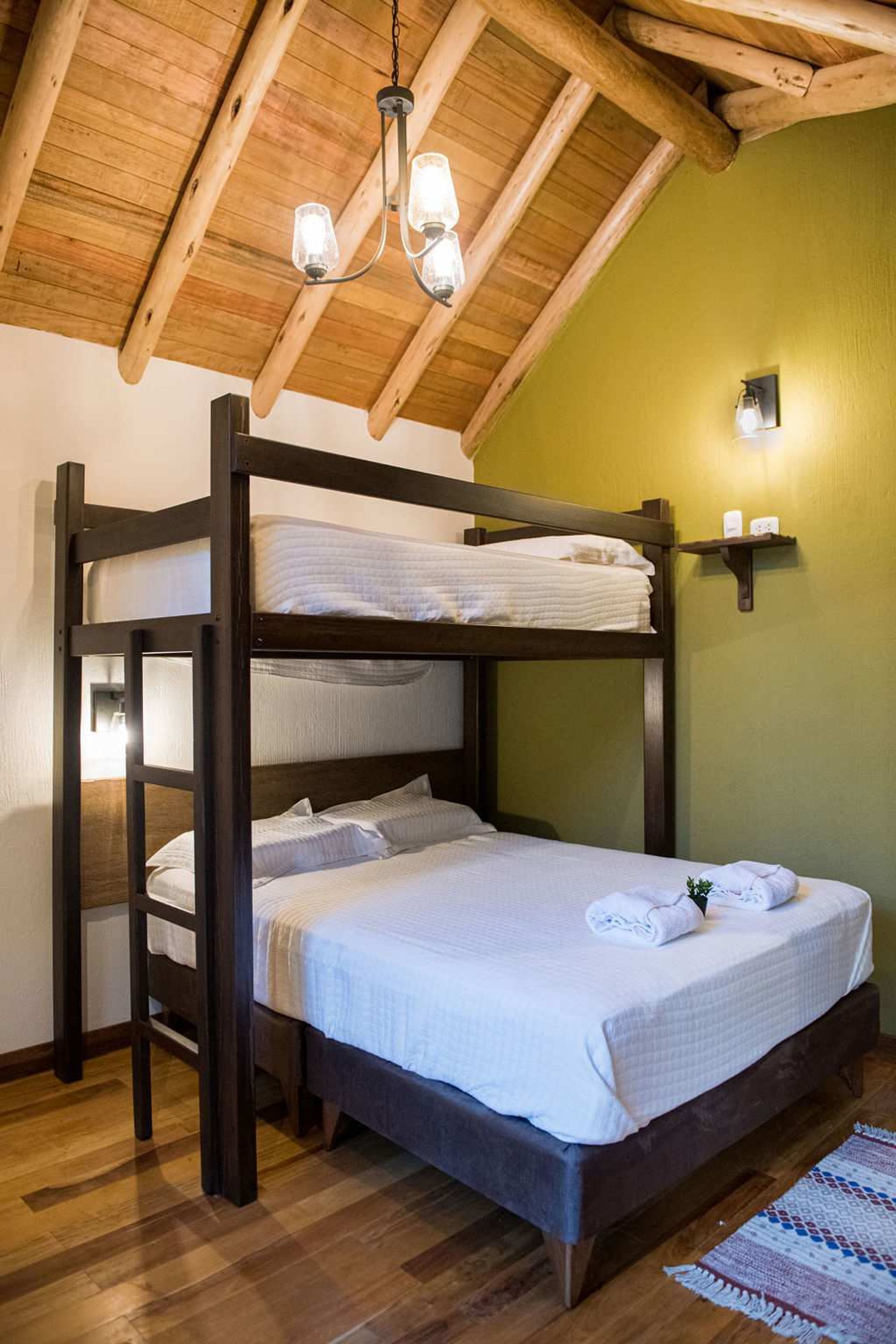 Quarto com beliche, vigas de madeira, lustre, luminária na parede e tapete estampado.