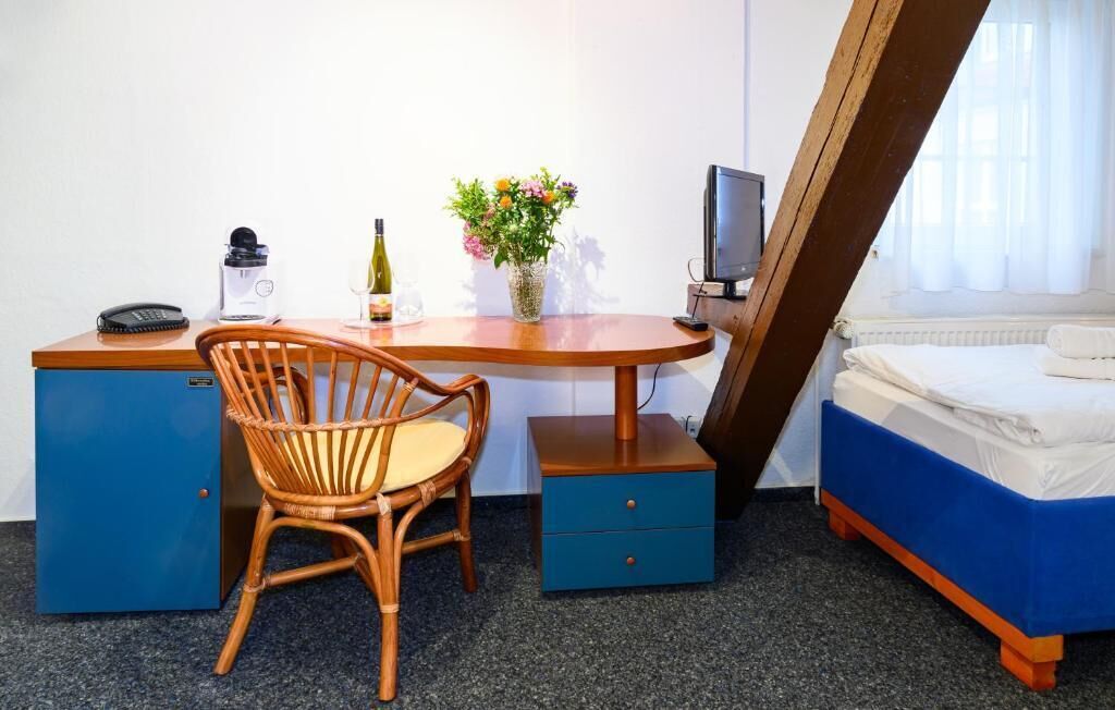 Quarto de hotel compacto com escrivaninha de madeira, cadeira de vime, mesa de cabeceira pequena, cama com colcha branca e azul, e televisão.