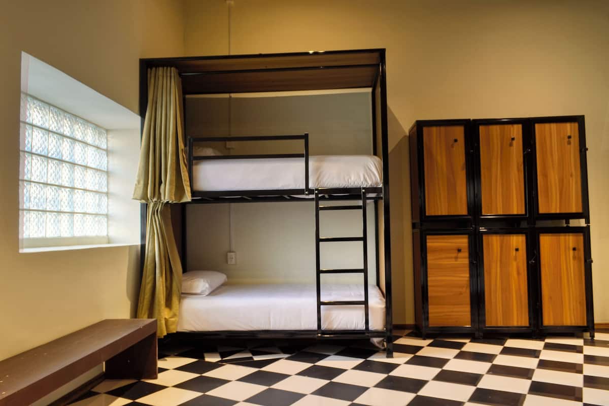Uma cama beliche com escada, um banco e um conjunto de armários de madeira num quarto com piso xadrez.