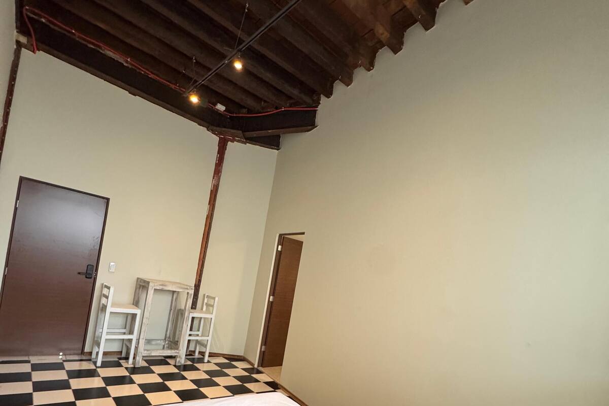 Quarto com piso xadrez, cama com roupa de cama branca, teto de madeira e uma porta.