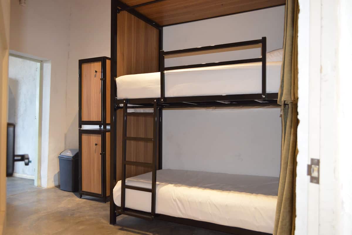Uma cama beliche com escada de madeira, um pequeno armário e uma lixeira em um quarto com parede lisa e uma porta ao fundo.