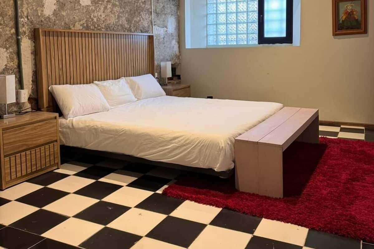 Quarto com chão xadrez, cama com cabeceira de madeira, janela com vista e tapete vermelho.