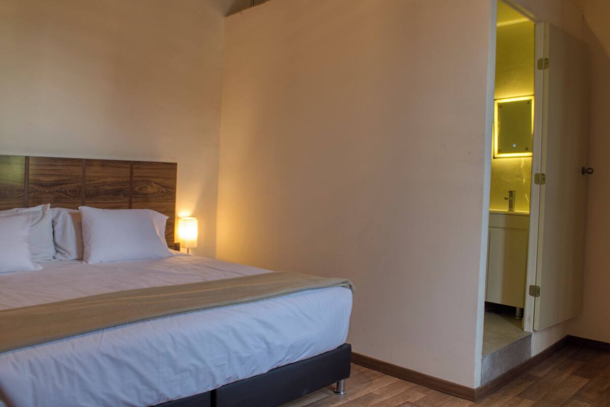 Quarto de hotel com cama, mesa de cabeceira e porta de acesso a um banheiro.