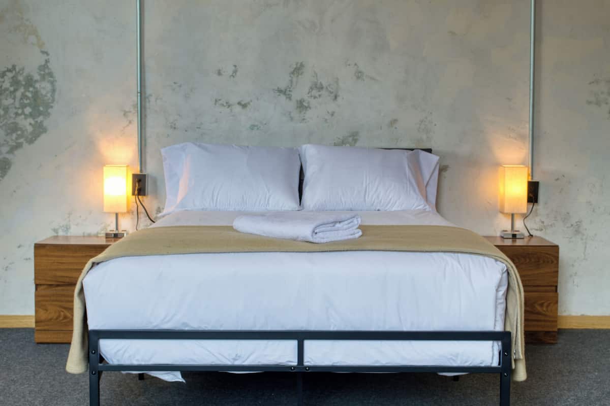 Uma cama bem arrumada com lençóis brancos e um cobertor bege, com duas mesas de cabeceira com luminárias, em um quarto com parede texturizada.