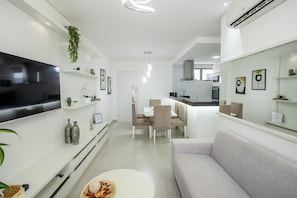Uma sala de estar moderna com televisão de tela plana, um sofá, uma área de jantar com mesa e cadeiras, e uma cozinha ao fundo.