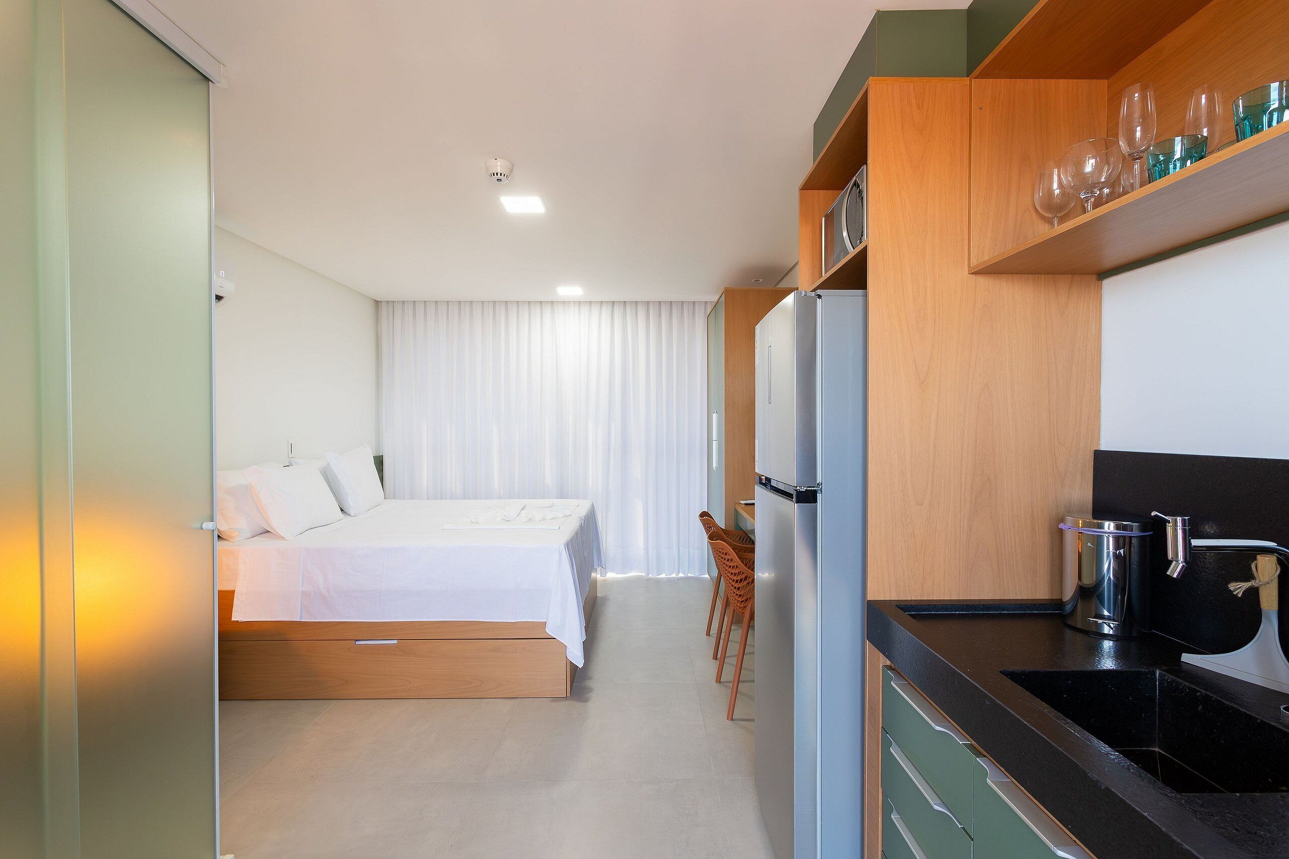 Quarto de hotel moderno com cama, área de cozinha com frigorífico e mesa de jantar.