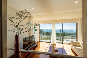 Uma sala de estar moderna com uma TV grande, uma mesa de centro de madeira, um sofá e vista para o exterior através de grandes janelas.