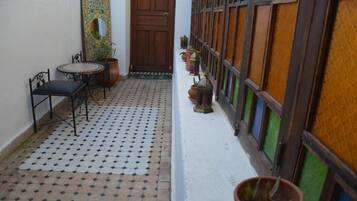 Um corredor estreito com chão de azulejos, porta de madeira, uma pequena mesa com uma cadeira e plantas em vasos ao longo da parede.